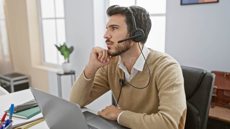 A Focused Hispanic Man with a Headset Using a Laptop in a Modern Office ...