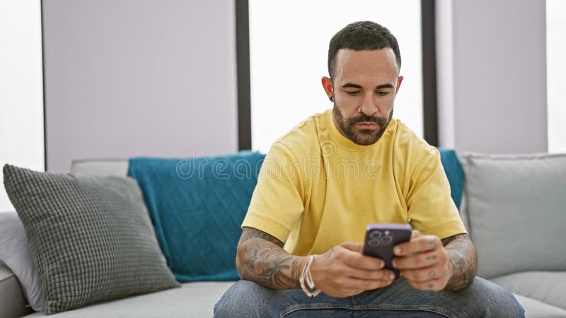 A Focused Hispanic Man with Tattoos Uses a Smartphone while Sitting on ...