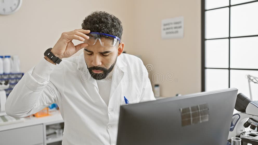 A Focused Hispanic Man in a White Lab Coat Examines Data on a Computer ...