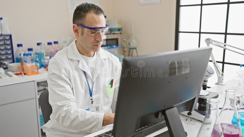 A Focused Hispanic Scientist in a Lab Coat and Safety Glasses Works on ...