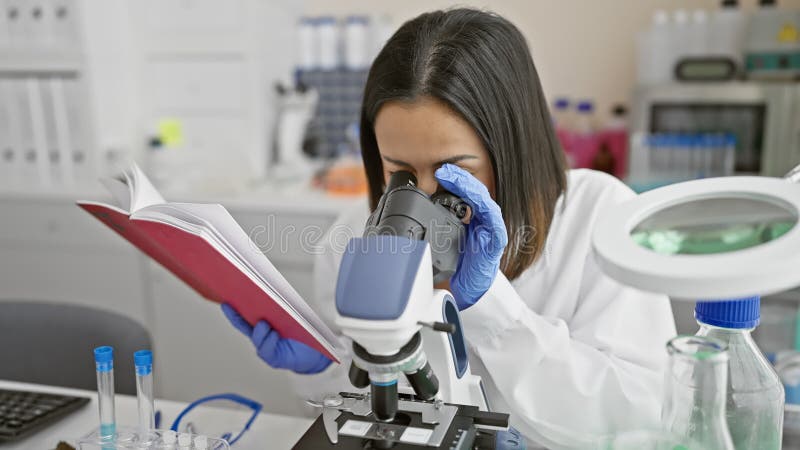 A Focused Latina Scientist Analyzes Samples Under a Microscope while ...