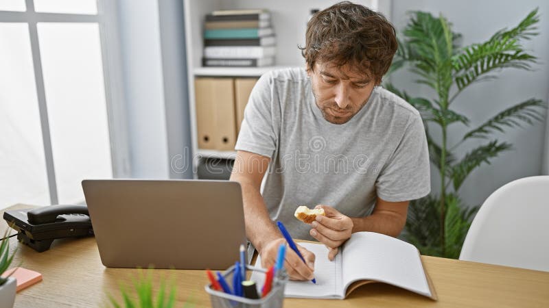 A Focused Man Snacking while Writing in a Notebook at His Modern Office ...