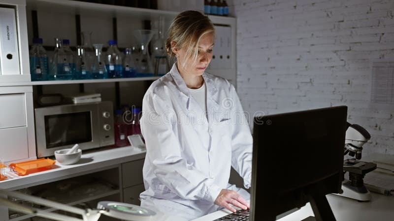 A Focused Woman Scientist in a Lab Coat Works on a Computer in a Modern ...