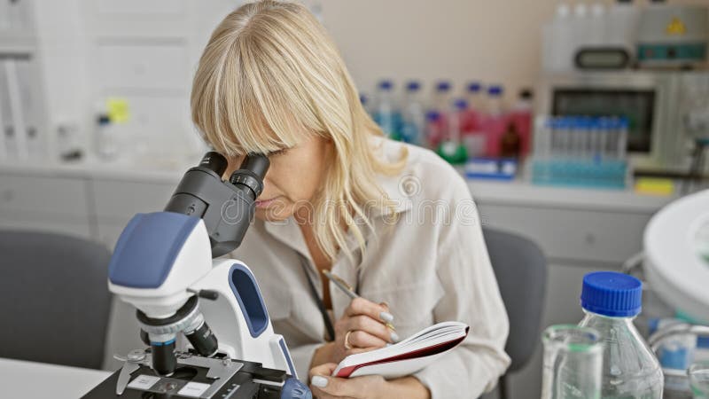 A Focused Woman Scientist Uses a Microscope and Takes Notes in a ... A Focused Woman Scientist Uses a Microscope and Takes Notes in a ...