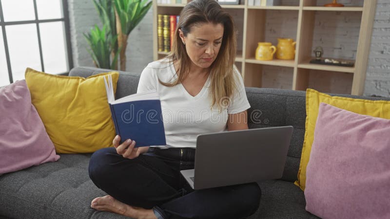 A Focused Woman Sits on a Couch at Home, Multitasking with a Laptop and ...