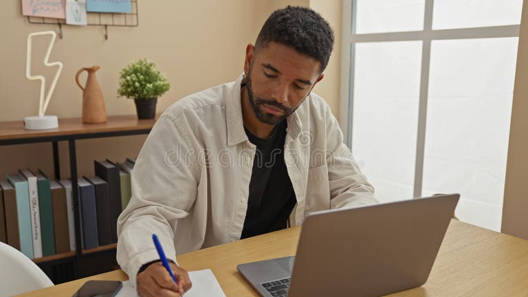 A Focused Young Black Man with a Beard Writing Notes and Using a Laptop ...
