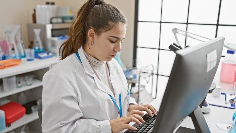 A Focused Young Hispanic Woman in a Lab Coat Typing on a Computer in a ...
