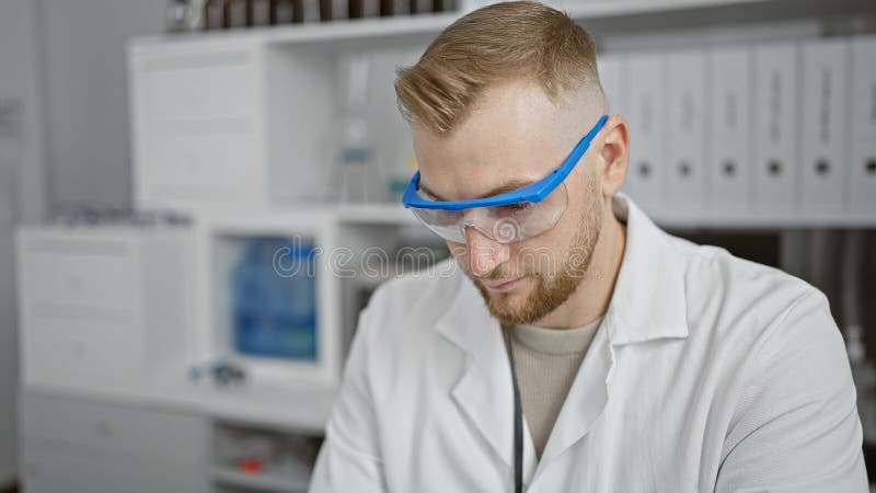 A Focused Young Man with a Beard Wearing Safety Goggles and a Lab Coat ...