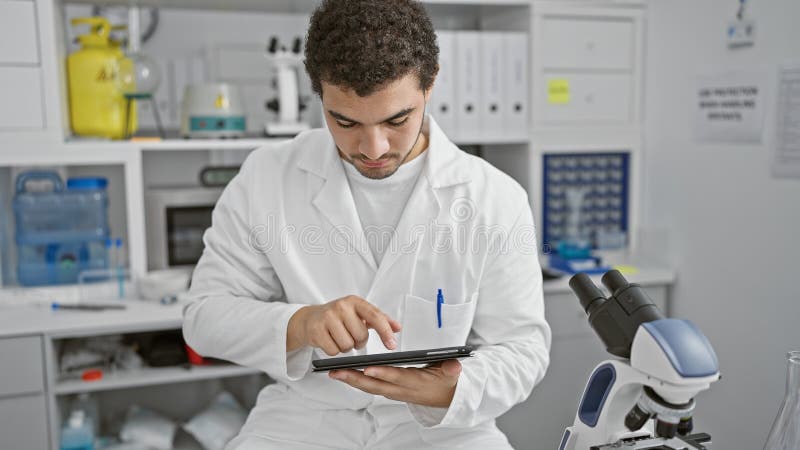 A Focused Young Man in a Lab Coat Studies Results on a Tablet in a ...