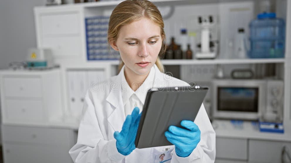 A Focused Young Woman in a Lab Coat and Gloves Using a Tablet in a ...