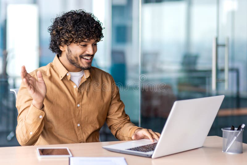 Friendly Middle Eastern Programmer Waving Hand while Using Portable Computer for Remote Online ...