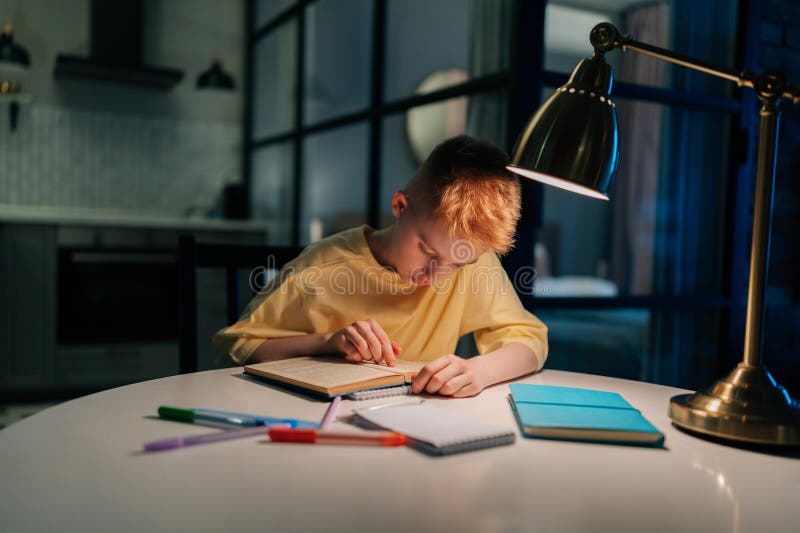 Side View of Focused Schoolboy Studying at Home Doing Homework Sitting at Table Under Light of ...