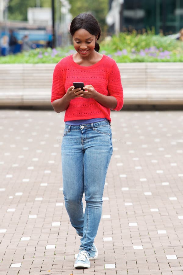 Full Length Smiling Young Black Woman Walking with Cellphone in the ... Full Length Smiling Young Black Woman Walking with Cellphone in the ...