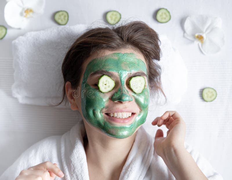 Funny Young Woman With A Cosmetic Mask On Her Face And Cucumbers On Her
