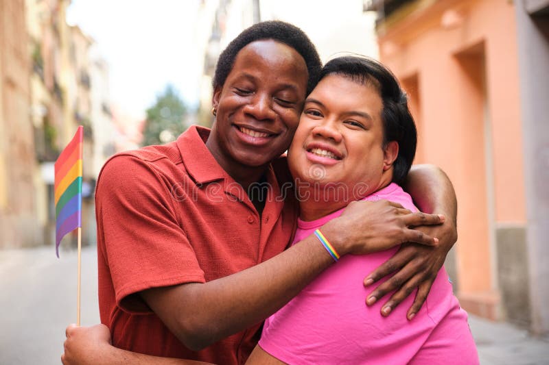 Gay Multiracial Couple Hug Each Other While One Of Them Holds A Rainbow