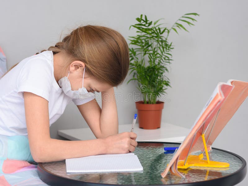 A Girl in a Medical Mask Does Homework at a Table in Front of the Bed ...