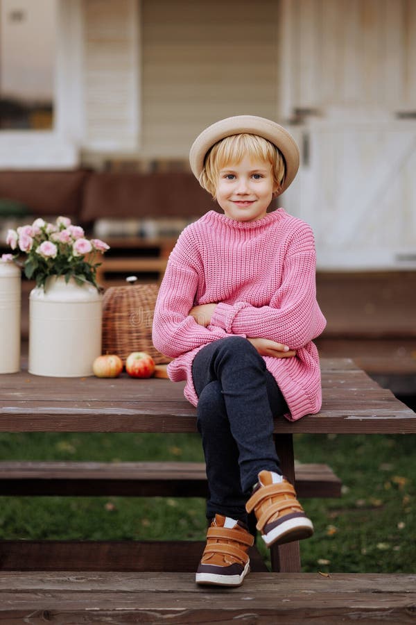 A Girl in a Pink Sweater and Hat Sits on a Wooden Table in the Backyard ...