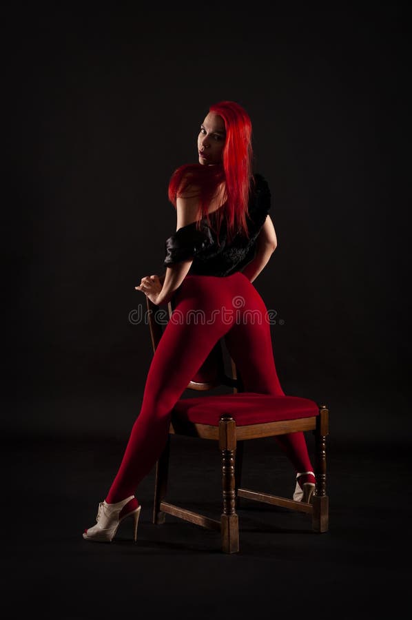 A Girl With Red Hair Wearing Red Leggings Poses On A Chair On A Dark