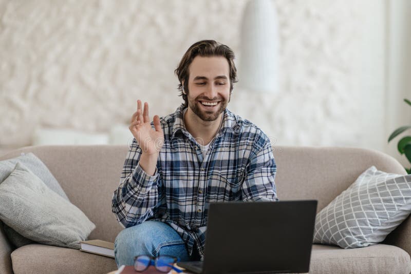 Glad Millennial Caucasian Guy With Stubble Sits On Sofa Waving Hand And