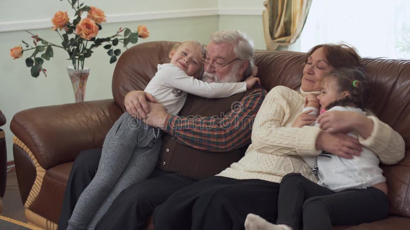 Grandfather And Grandmother Sitting On Leather Sofa In Modern Living Room With Two Small Girls