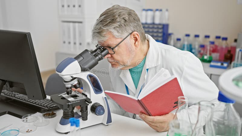 Grey-haired Man in Lab Coat Using Microscope and Holding a Notebook in ...