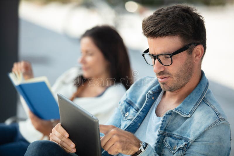 Guy Uses Tablet Next To Sitting Girl Reading Book Stock Image - Image ...
