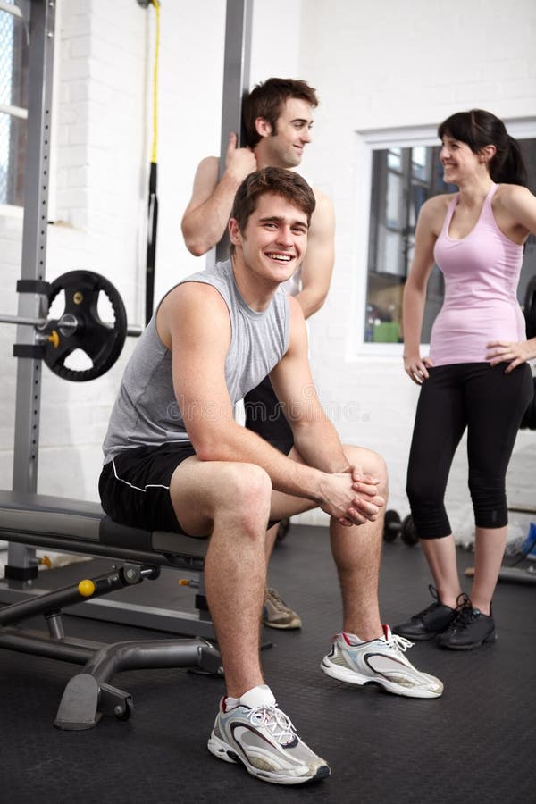 Gym Buddies Young Man Smiling At The Camera With Two Other Gym Members Chatting Behind Him