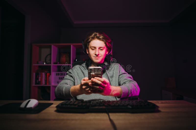 Handsome Guy is Sitting at the Computer at Night and Uses a Smartphone ...