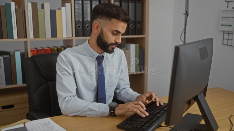 Handsome Hispanic Man with Beard Working at a Desk in an Office Setting with Bookshelves and ...