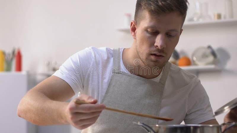 Handsome Man in Apron Cooking, Stirring Ingredients in Pan and Trying ... Handsome Man in Apron Cooking, Stirring Ingredients in Pan and Trying ...