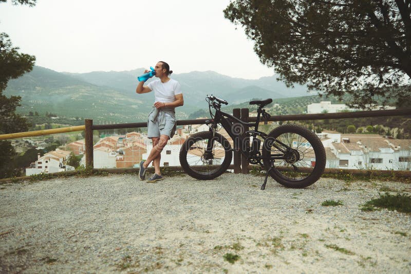 Handsome Muscular Man Cyclist Drinks Water From Sports Bottle Standing By Wooden Fence Next To