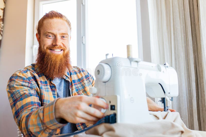 Handsome Redhaired Man with Long Beard Sews at a Sewing Machine at Home ...