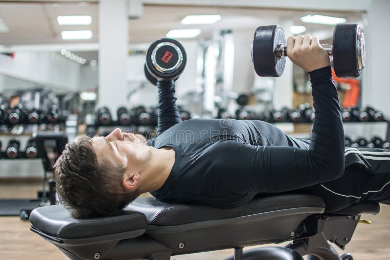 Handsome Young Man Lying on Exercise Bench and Lifting Dumbbells at Gym ...