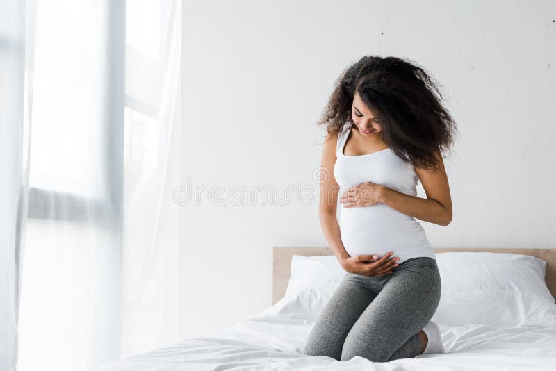 African American Pregnant Woman Touching Belly While Sitting On Bed