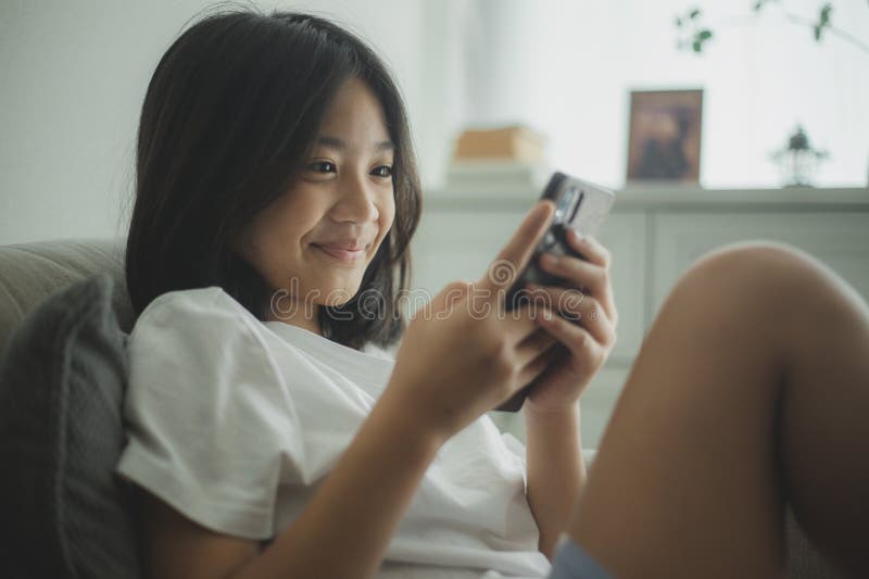 Happy Asian Girl Relaxing On A Comfortable Couch At Home Texting And Messaging On A Smartphone