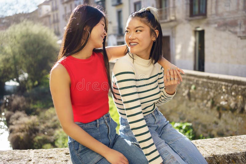Happy Asian Girlfriends Sitting Together On Street And Looking At Each