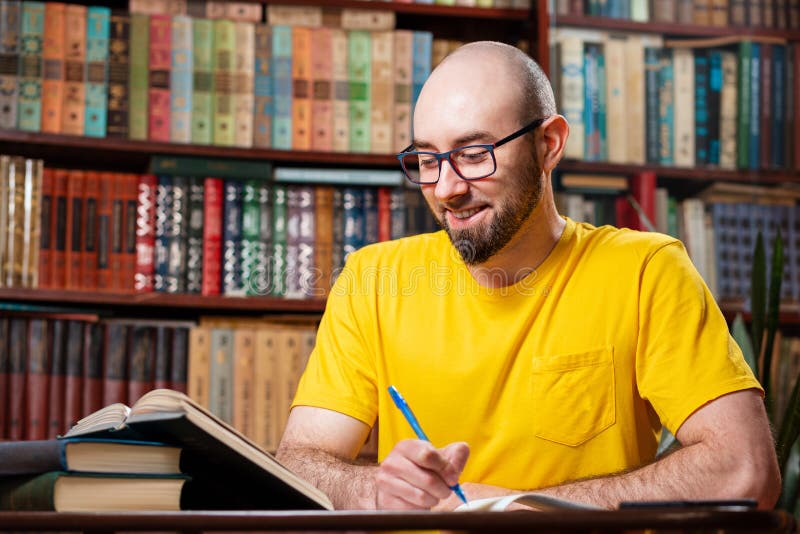 A Happy Bearded Bald Man With Glasses Uses A Smartphone Bookshelves In