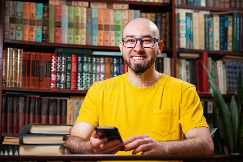 A Happy Bearded Bald Man With Glasses Uses A Smartphone Bookshelves In