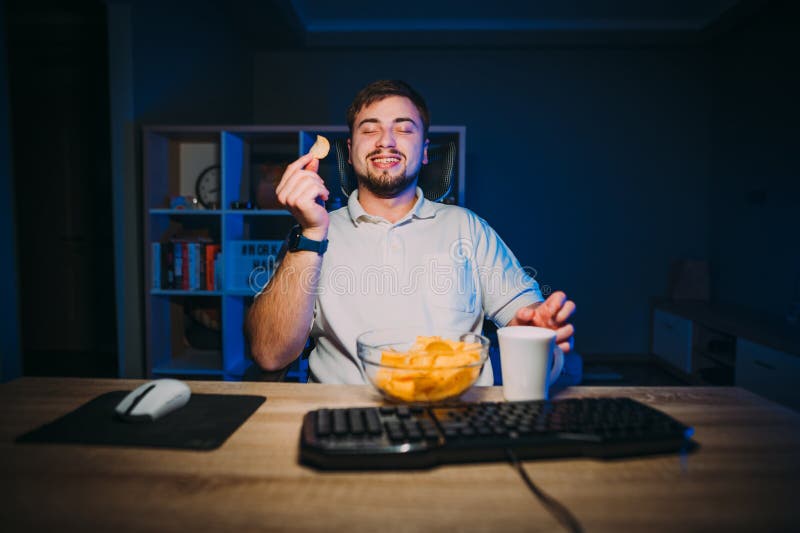 Happy Bearded Man Sits at Work at the Computer at Night and Eats Chips ...