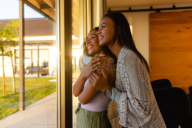 Happy Biracial Lesbian Couple Embracing And Looking Out Window At Home
