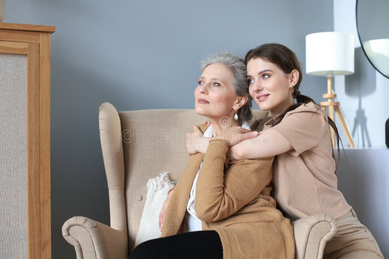 Happy Daughter Hugging Older Mother Standing Behind Chair In Living
