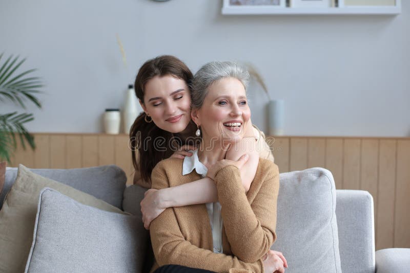 Happy Daughter Hugging Older Mother Standing Behind Chair In Living