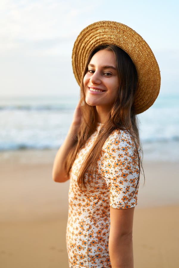 Smiling Young Woman In Floral Dress And Straw Hat On Sandy Beach At