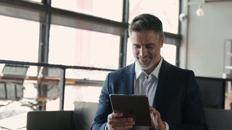 Happy Mature Business Man Wearing Suit Standing In Office Using Digital