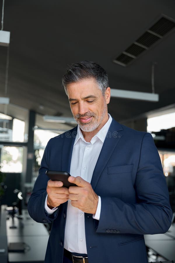 Happy Mature Business Man Wearing Suit Standing In Office Using Mobile