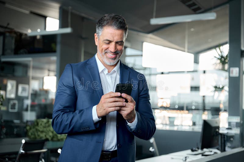 Happy Mature Business Man Wearing Suit Standing In Office Using Mobile