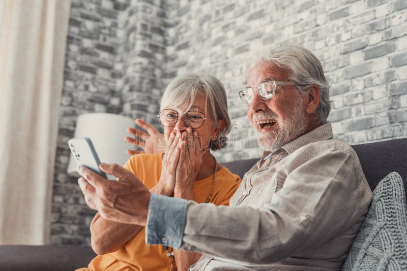 Happy Older Couple Sit On Couch Staring At Cellphone Screen Gesturing