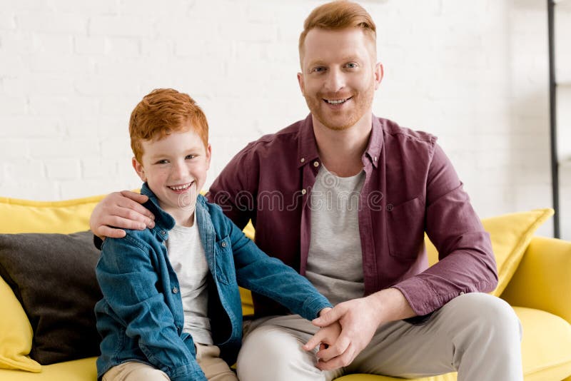 Happy Redhead Father And Son Smiling At Camera While Sitting Together