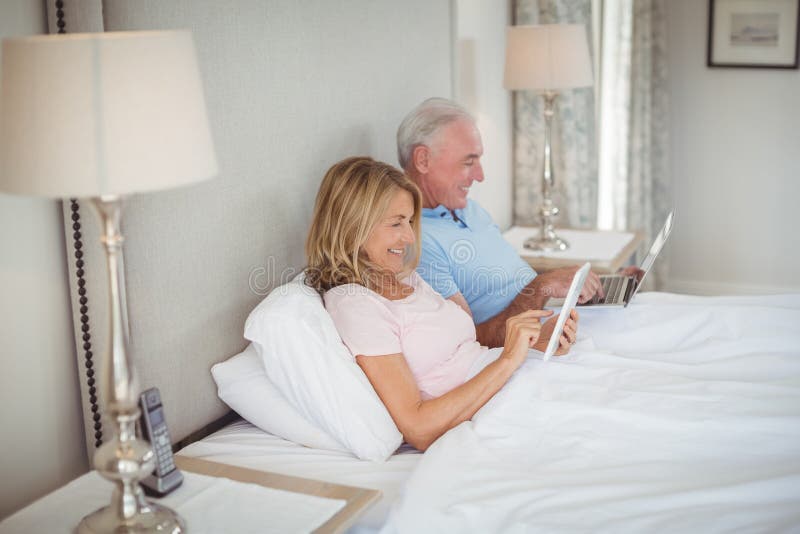 Happy Senior Couple On Bed Using Laptop And Digital Tablet Stock Image