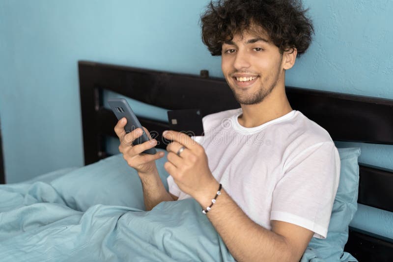 Happy Smiling Guy Using Mobile Phone and Credit Card at Home in Bed ...